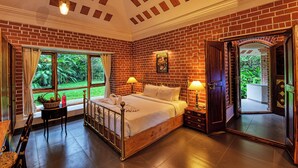 Um quarto com parede de tijolos, uma janela grande, uma cama, mesa de cabeceira e porta que dá para o exterior.