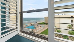 Vista pela janela de uma área de piscina com várias piscinas, guarda-sóis e uma praia ao fundo.