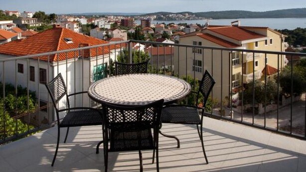 Uma varanda com uma mesa redonda e cadeiras, dando vista para uma cidade costeira com edifícios de telhado vermelho e uma extensão de água.