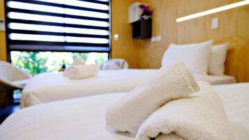 Quarto de hotel com duas camas, cada uma com uma toalha enrolada. Há uma parede de madeira e uma janela com venezianas.