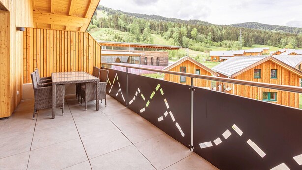 Varanda de madeira com mesa e cadeiras, com vista para uma fileira de cabanas em uma região montanhosa.