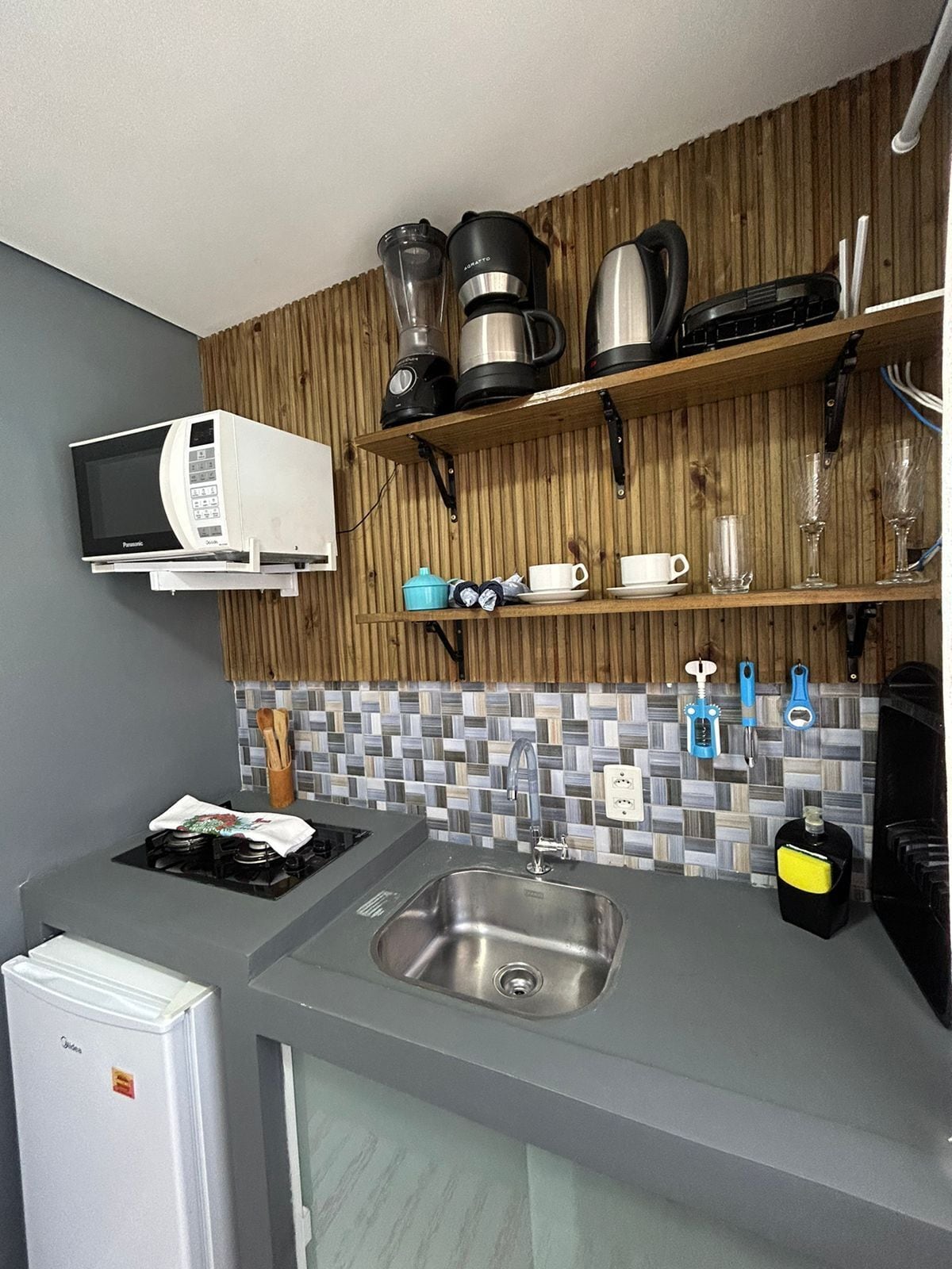 Uma cozinha compacta com micro-ondas, chaleira e máquina de café em prateleiras de madeira, pia com torneira e painel de azulejos.