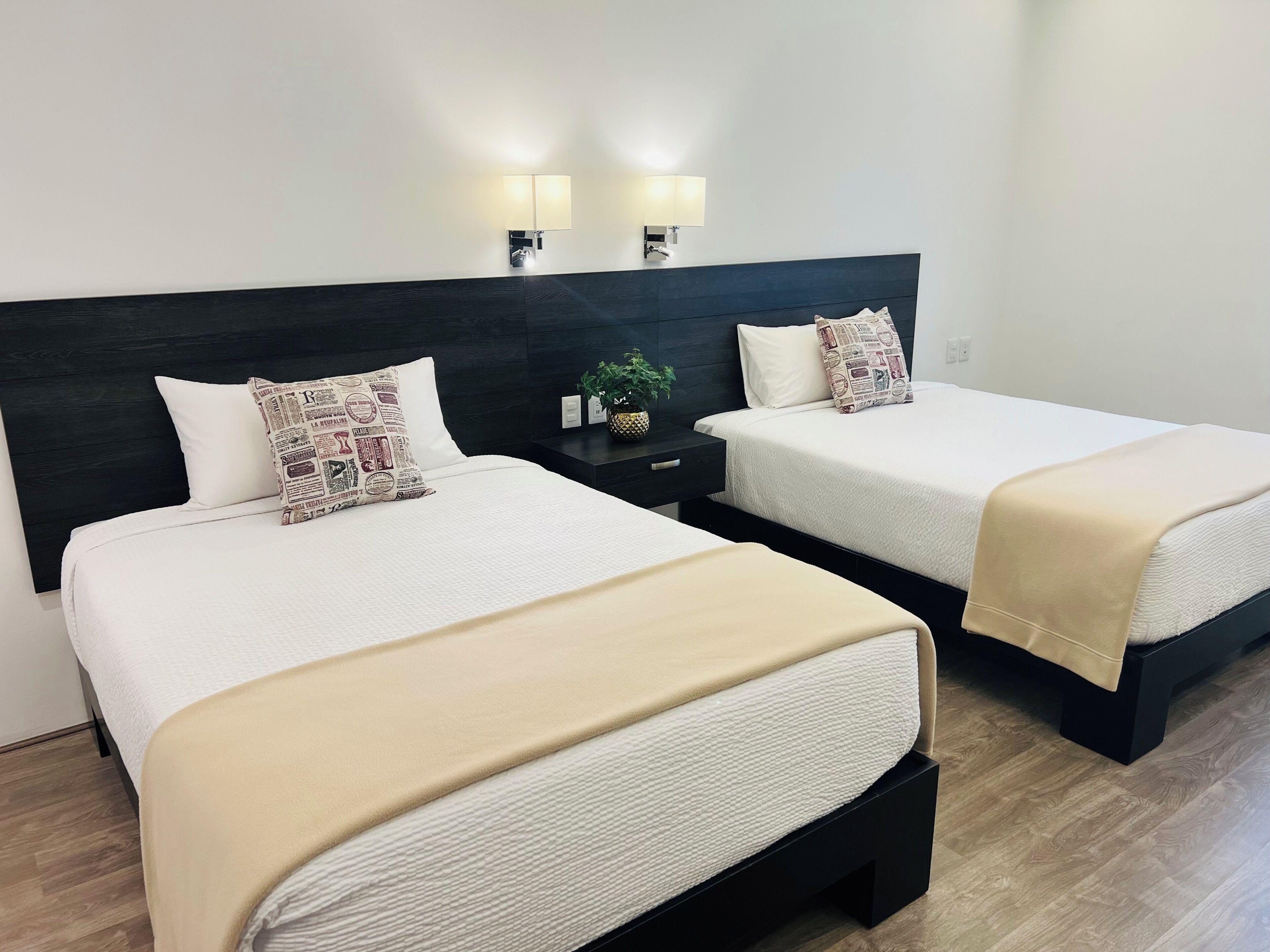 Duas camas com roupa de cama branca e mantas bege, uma cabeceira escura e duas luzes fixadas na parede.