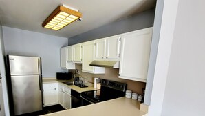 Cozinha compacta com armários brancos, geladeira de aço inoxidável, fogão preto e uma cadeira de madeira.