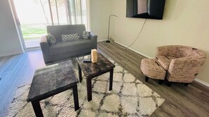 Uma sala de estar com sofá, duas mesinhas de centro, tapete estampado, televisão montada na parede e poltrona estampada.