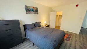 Quarto com cama, cómoda, mesa de cabeceira, abajur e um quadro na parede.