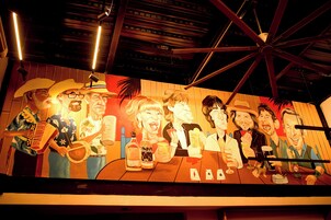 Um mural retrata pessoas jogando cartas e bebendo, com uma mesa e cadeiras de madeira.