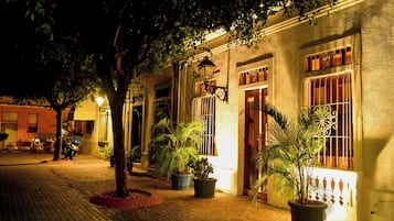 Uma cena noturna de rua com um edifício de fachada azulejada, várias janelas com grades de metal e plantas em vasos.