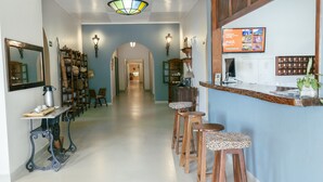 Um hall de hotel com um corredor longo, banquetas de madeira, um balcão de recepção e uma TV na parede exibindo um anúncio da PIRA.