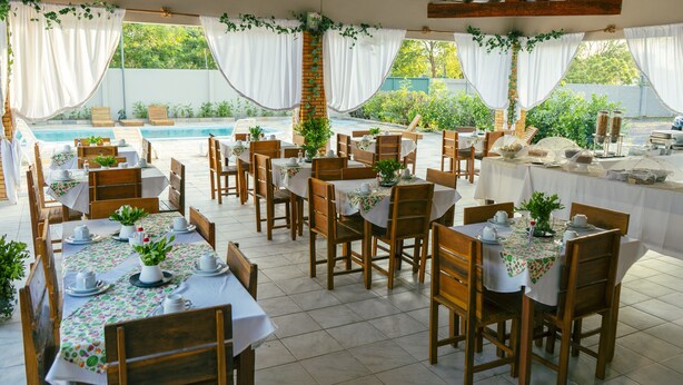 Uma área de refeições ao ar livre com mesas preparadas para uma refeição, rodeada por cadeiras, e uma piscina ao fundo.