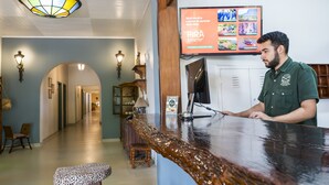 Uma área de recepção com balcão de madeira, uma televisão exibindo um menu e um corredor com um abajur e um banco.