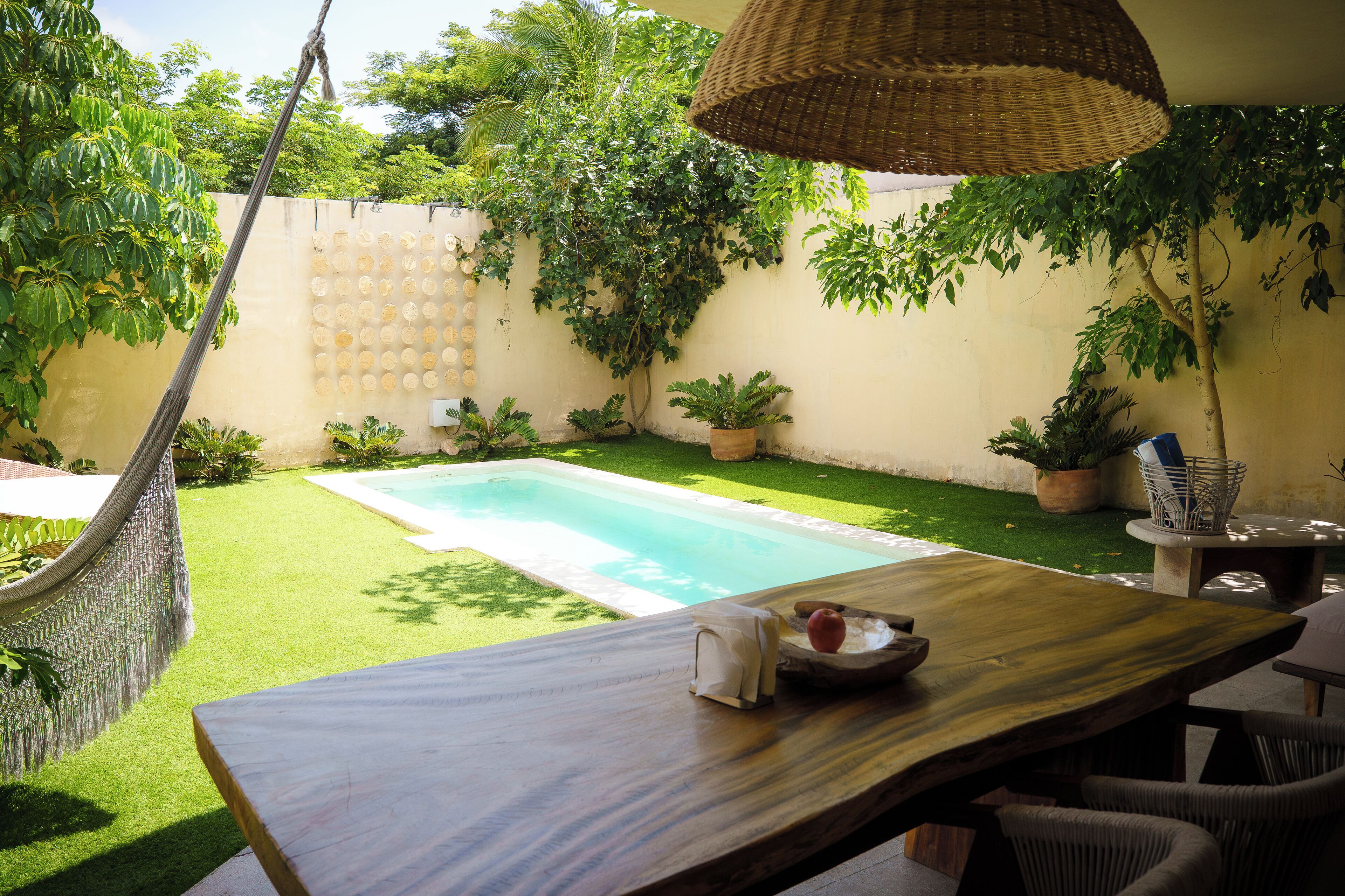Uma mesa de madeira com uma tigela e guardanapo, uma rede pendurada, uma piscina pequena e plantas em vasos num quintal.