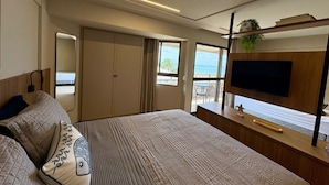 Quarto de hotel com cama, televisão, escrivaninha e vista para o oceano.