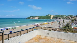 Uma praia com guarda-sóis, um caminho de azulejos e vista para o oceano e as colinas.