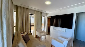 Uma sala de estar moderna com um sofá, duas poltronas, uma TV de tela plana na parede e grandes janelas com cortinas.