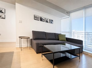Uma sala de estar moderna com um sofá cinzento, uma mesa de centro de vidro e uma pequena mesa lateral. Há quadros nas paredes e uma janela ampla com vista para a cidade.