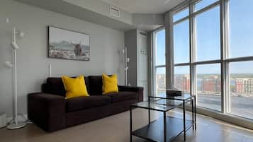 Uma sala de estar moderna com um sofá marrom escuro, dois travesseiros amarelos, uma mesa de centro de vidro e uma grande janela com vista para a cidade.
