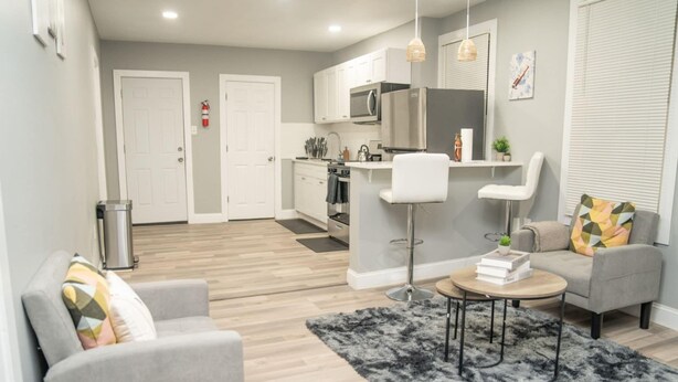 Uma sala de estar moderna com um sofá cinzento, uma mesa de centro e um tapete. Há uma cozinha com micro-ondas e frigorífico.