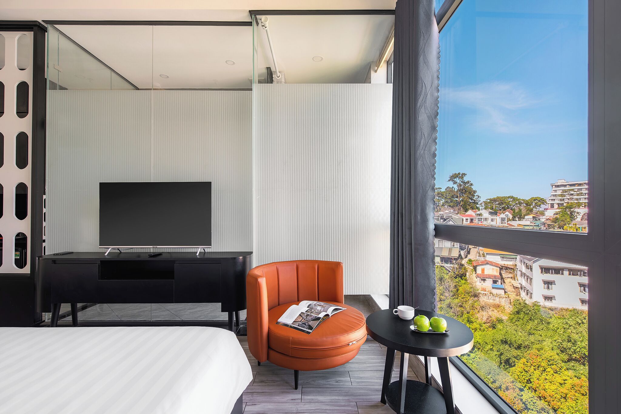 Quarto de hotel moderno com uma cama, uma televisão em um suporte preto, uma poltrona laranja, uma pequena mesa com uma revista e duas maçãs, e uma grande janela com vista para a cidade.