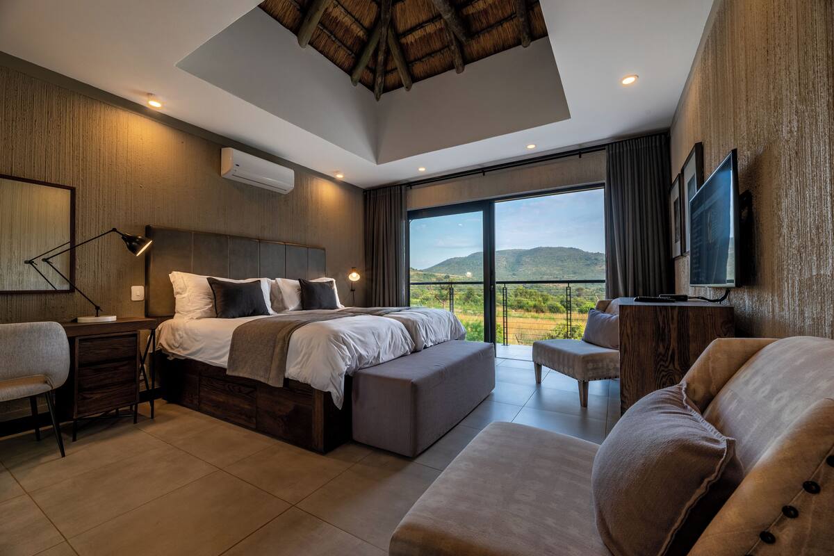 Quarto moderno com cama grande, escrivaninha, cadeira, televisão e varanda com vista para as colinas.
