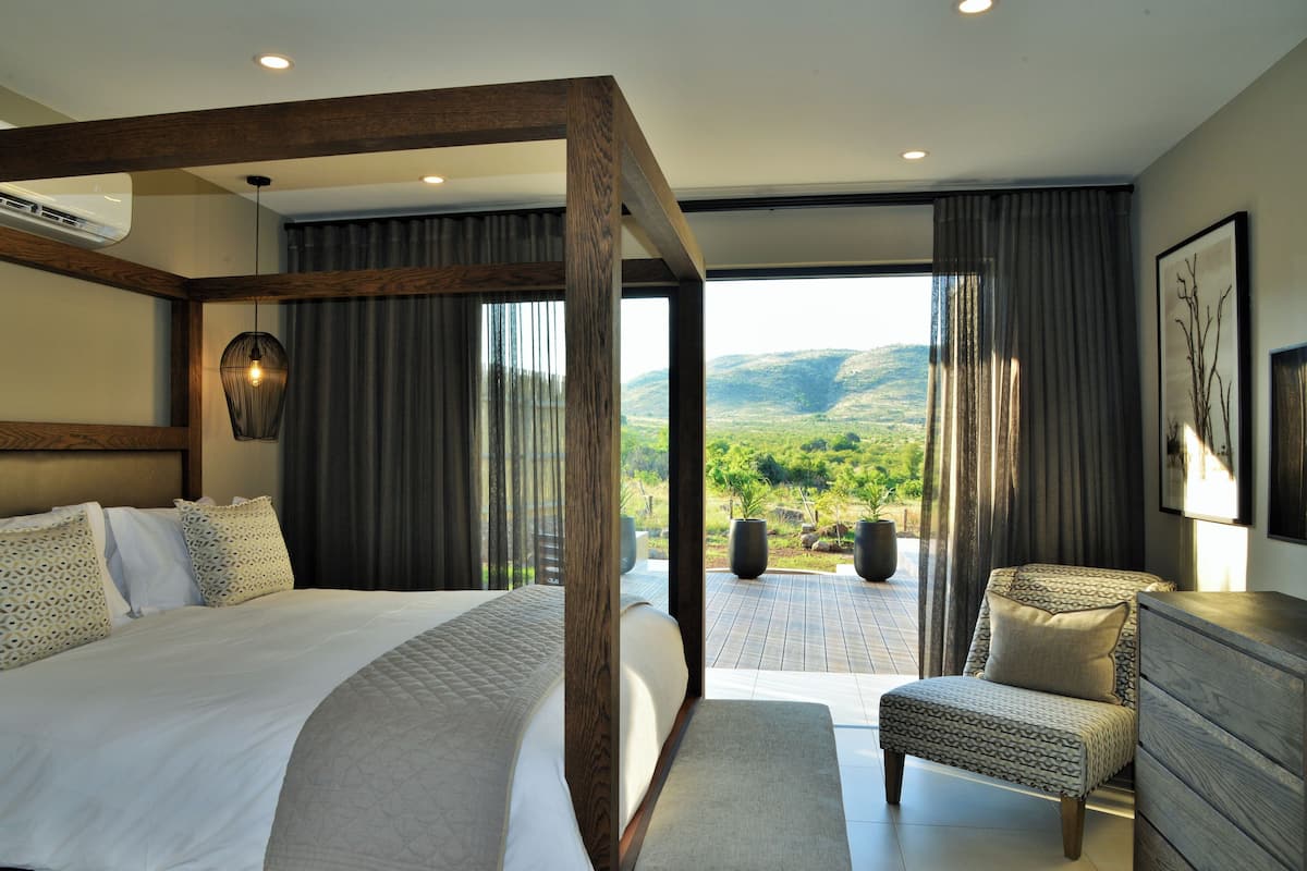 Quarto com cama de dossel, vista para o exterior e área de estar com poltrona e cômoda.