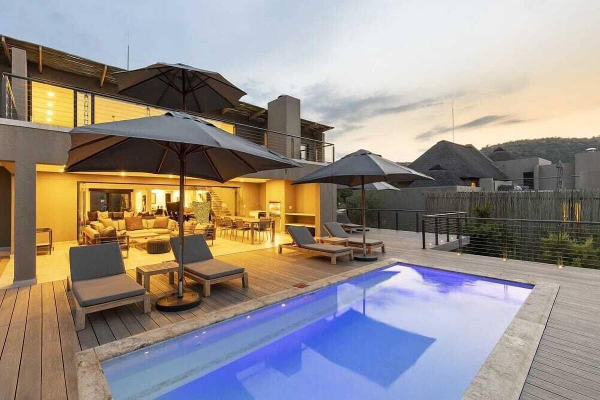 Uma área moderna de piscina externa com espreguiçadeiras e guarda-sóis, um deck de madeira e um interior bem iluminado com área de estar.