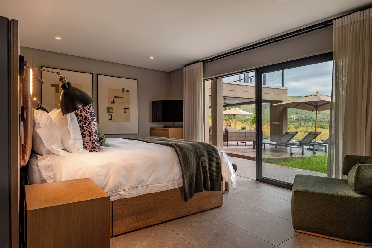 Quarto moderno com cama grande, televisão e vista para um deck com mobiliário de exterior.
