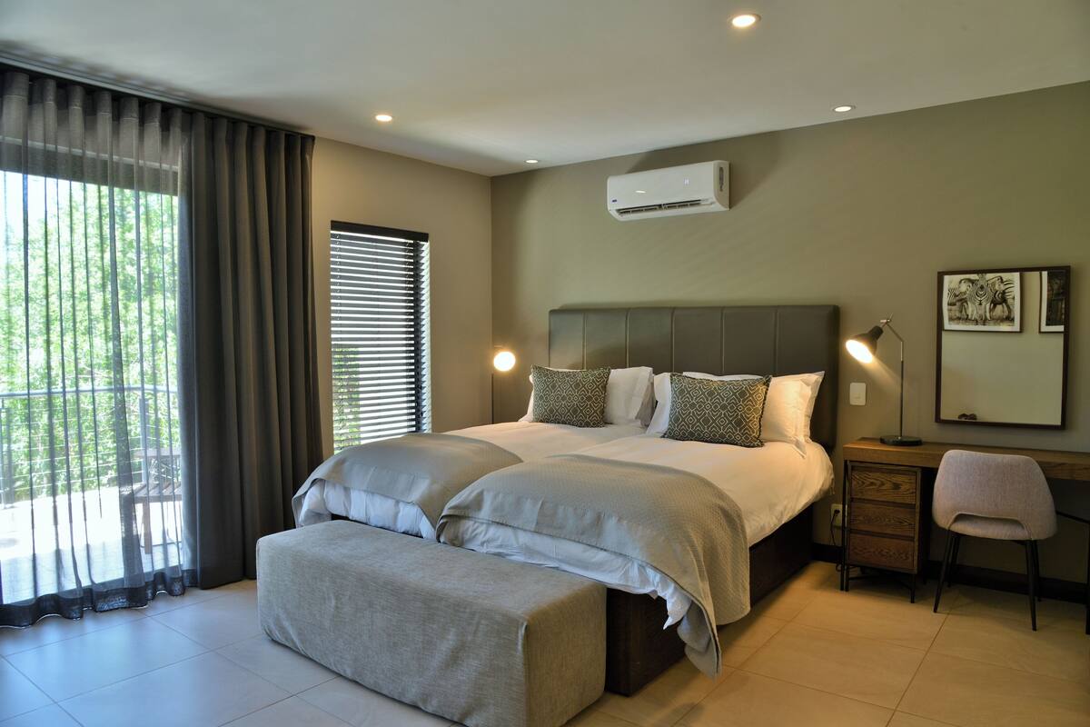 Quarto moderno com cama grande, mesas de cabeceira, aparelho de ar condicionado e vista para o verde pela janela.