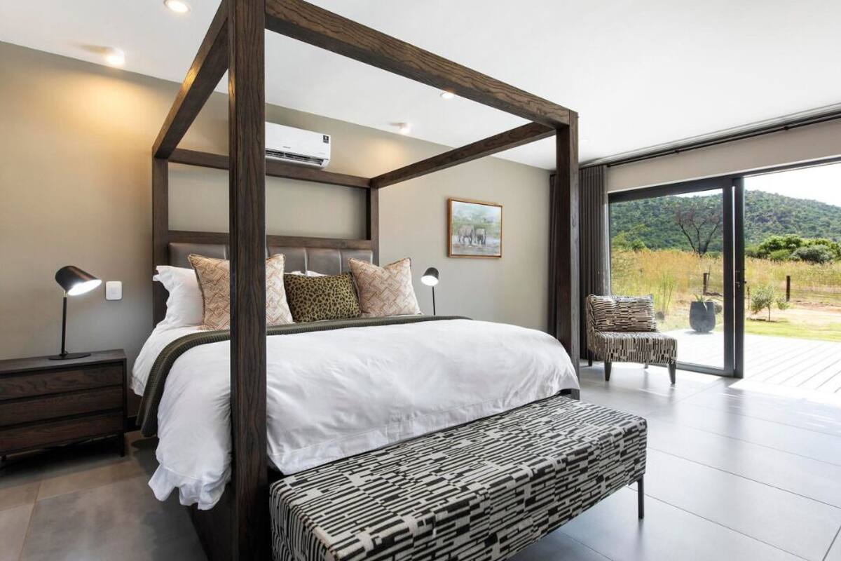 Quarto moderno com cama de dossel, um banco, mesa de cabeceira, abajur e vista para um jardim através de janelas amplas.