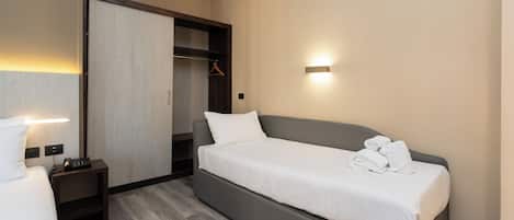 Quarto de hotel moderno com uma cama de solteiro, mesa de cabeceira com luminária, guarda-roupa e iluminação embutida na parede.