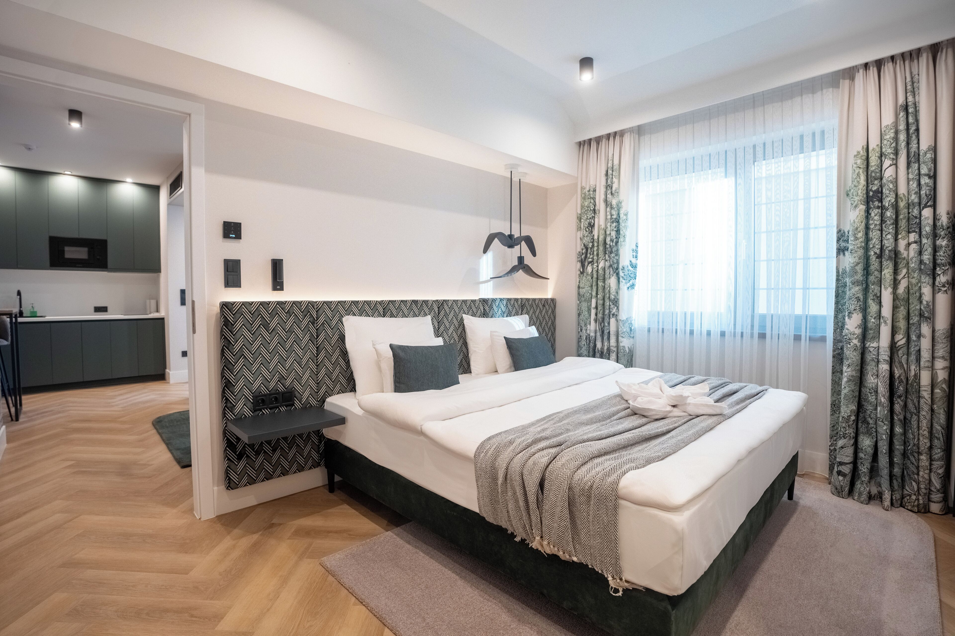 Quarto moderno com cama grande, mesa de cabeceira, guarda-roupa embutido, pequena cozinha e uma janela ampla com cortinas.
