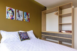 Quarto com cama, roupa de cama branca, almofada colorida e três quadros na parede.