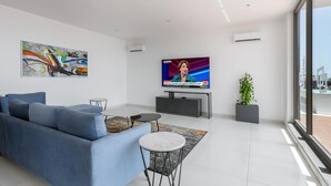 Uma sala de estar moderna com um sofá azul, uma mesa redonda pequena, uma televisão de tela plana e um grande quadro abstrato na parede.
