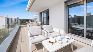 Uma varanda moderna com mobiliário branco, uma mesa com um livro e copos, e vista para a paisagem urbana.