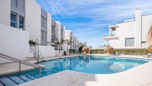 Uma área de piscina moderna com céu azul claro, edifícios brancos e algumas palmeiras.