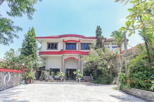 Um edifício de dois andares com telhado vermelho e colunas, rodeado por vegetação e plantas em vasos.