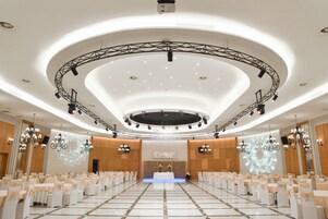 Uma sala de conferências ampla e bem iluminada, com palco, fileiras de mesas e cadeiras brancas, lustres e um design moderno no teto.