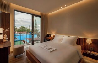 Quarto de hotel com uma cama grande, uma mesa, uma cadeira, uma mesinha de cabeceira, uma luminária e vista para uma piscina.