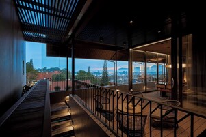 Uma varanda com vista para a cidade, com guarda-corpo de madeira, área de estar com cadeiras e escada que leva ao nível superior.
