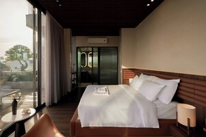 Quarto de hotel moderno com uma cama grande, uma mesa de cabeceira, uma luminária e vista para o exterior através de uma janela ampla.