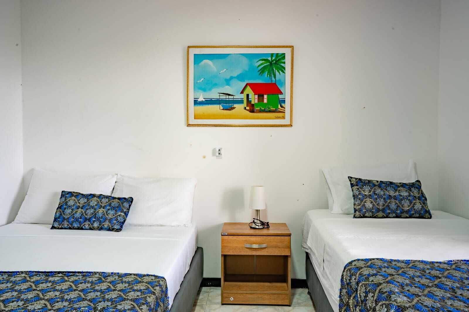 Quarto de hotel com duas camas, cada uma com colcha estampada, mesa de cabeceira com abajur e pintura de uma paisagem de praia na parede.