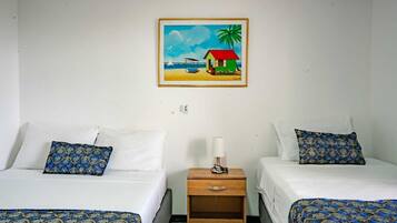 Quarto de hotel com duas camas, cada uma com colcha estampada, mesa de cabeceira com abajur e pintura de uma paisagem de praia na parede.