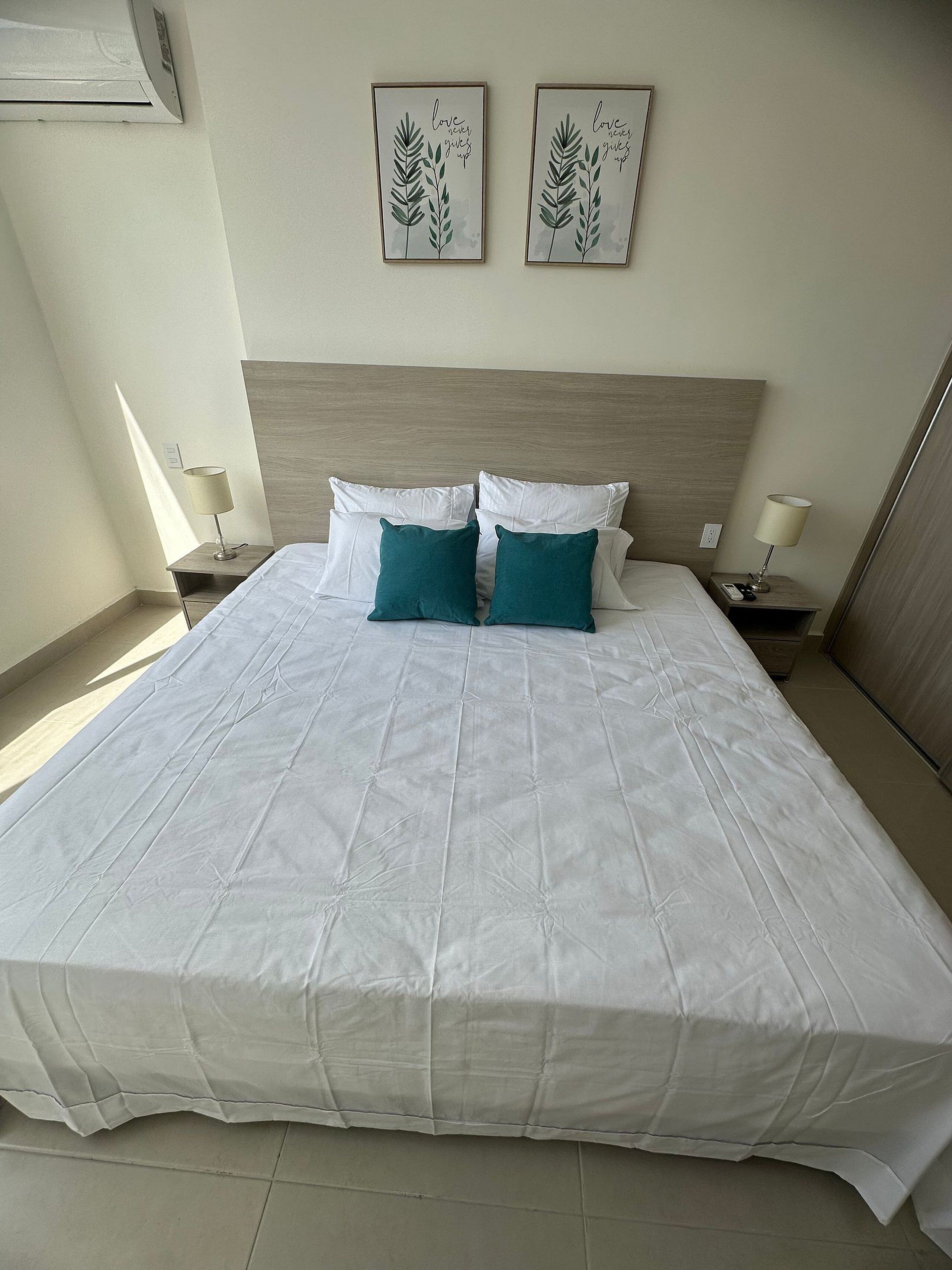 Cama bem arrumada com roupa de cama branca e dois travesseiros verde-água, uma cabeceira de madeira e duas obras de arte emolduradas acima.