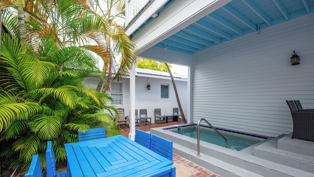 Uma área de piscina com mesa e cadeiras azuis, cercada por palmeiras e um edifício branco.