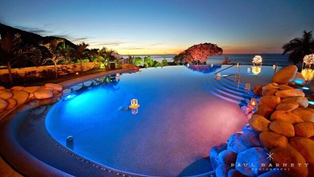 Uma grande piscina infinita com vista para o oceano ao pôr do sol, cercada por pedras e palmeiras.