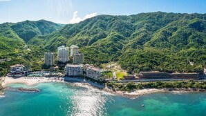 Vista aérea de um resort costeiro com vários edifícios, uma praia e colinas verdes exuberantes ao fundo.