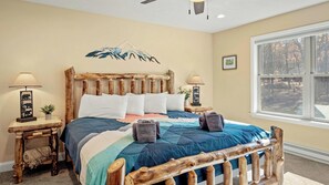 Quarto com cabeceira de madeira, cama com colcha azul e branca, duas mesas de cabeceira com abajures, janela com vista para árvores e uma obra de arte pendurada na parede.