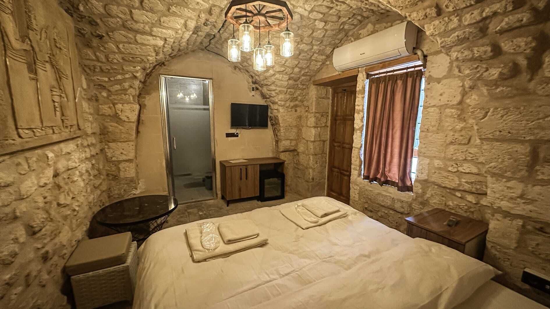 Quarto com paredes de pedra, uma cama, uma escrivaninha pequena, um espelho e uma janela com cortinas.