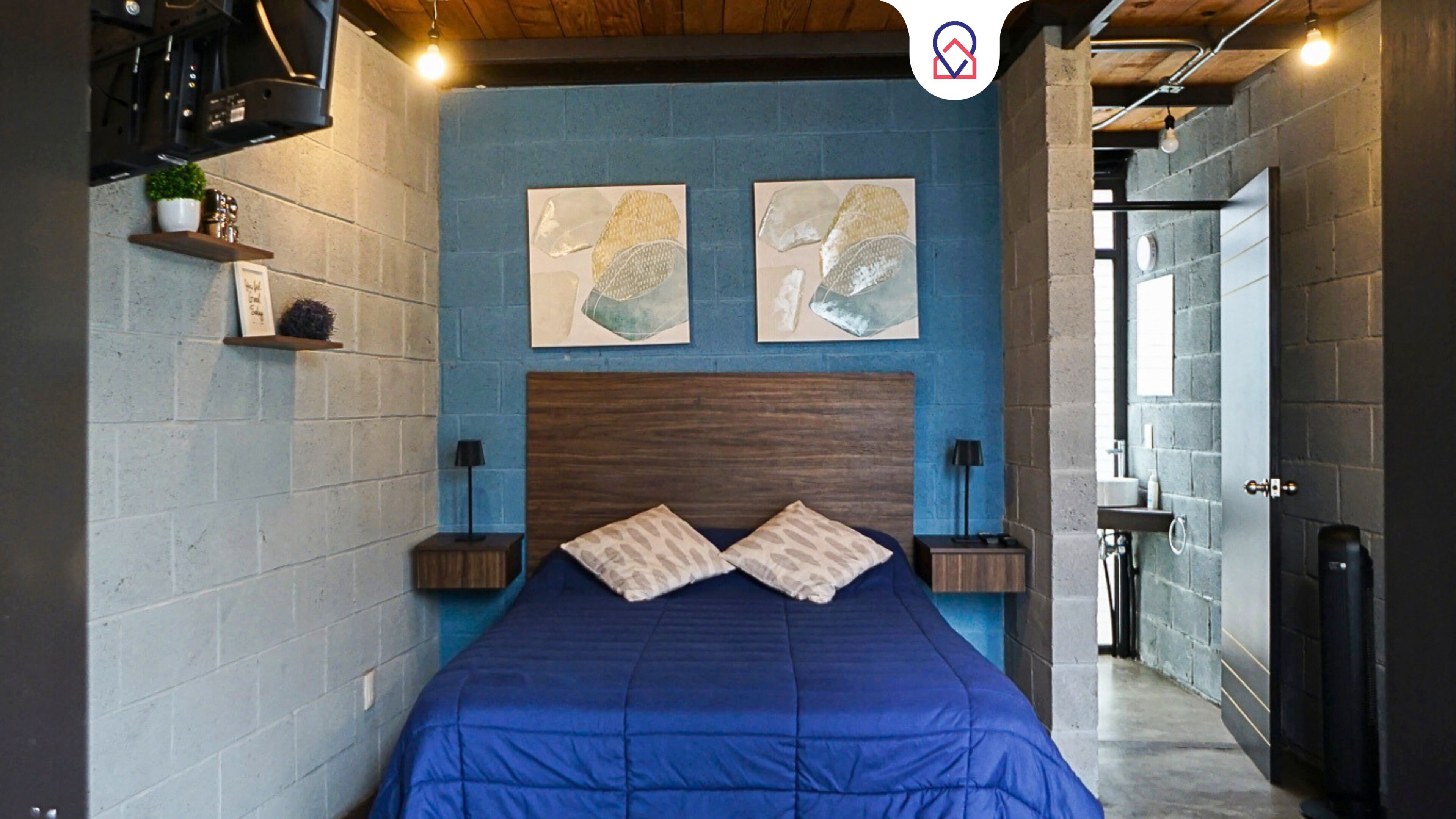 Quarto com colcha azul, duas pinturas penduradas na parede, cabeceira de madeira, mesas de cabeceira com abajures e parede de tijolos.