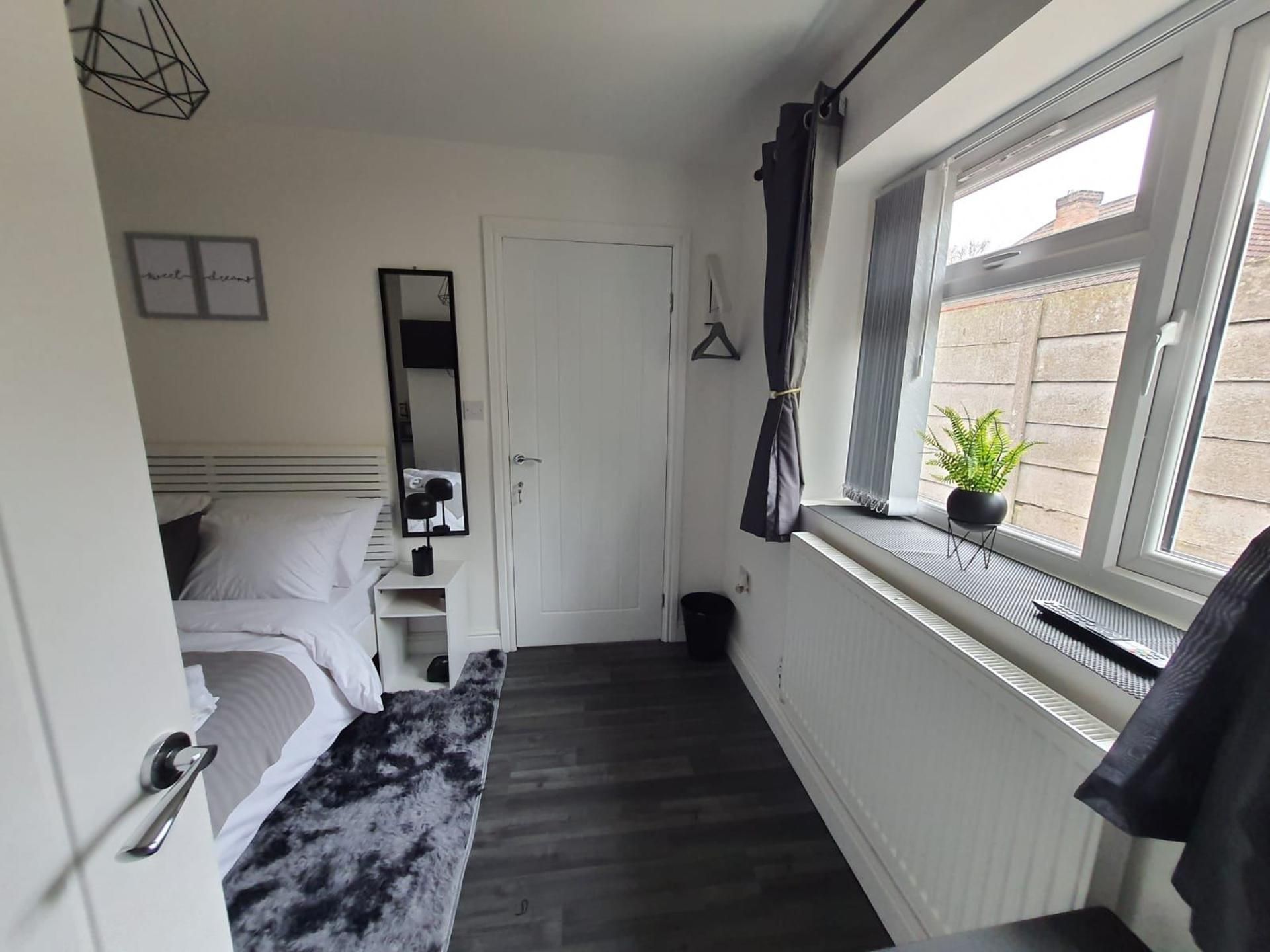 A narrow hotel room with a bed, a bench, a mirror, a radiator, and a window with a plant.